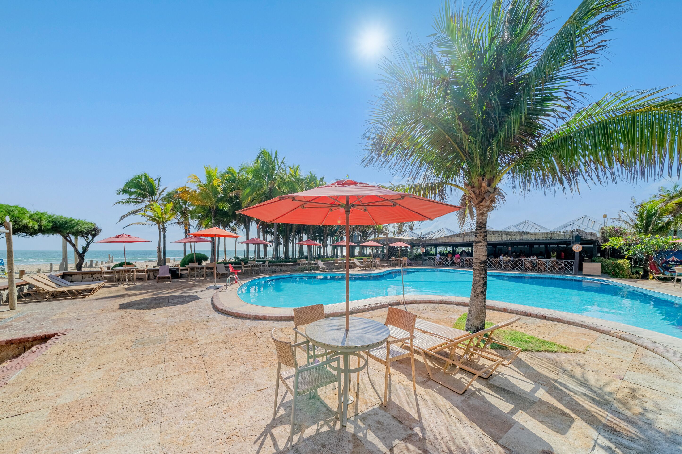 Área de piscina com espreguiçadeiras, guarda-sóis vermelhos e uma palmeira.