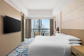 Quarto de hotel com uma cama grande, uma televisão de tela plana, uma mesinha com uma planta e vista para prédios altos através de amplas janelas.