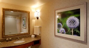 Uma casa de banho com bancada de granito, lavatório, espelho e um quadro com flores na parede.