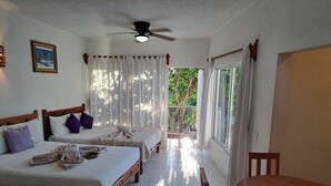 Quarto de hotel com duas camas, um ventilador de teto, janela com cortinas, uma mesa de madeira e vista para o verde lá fora.