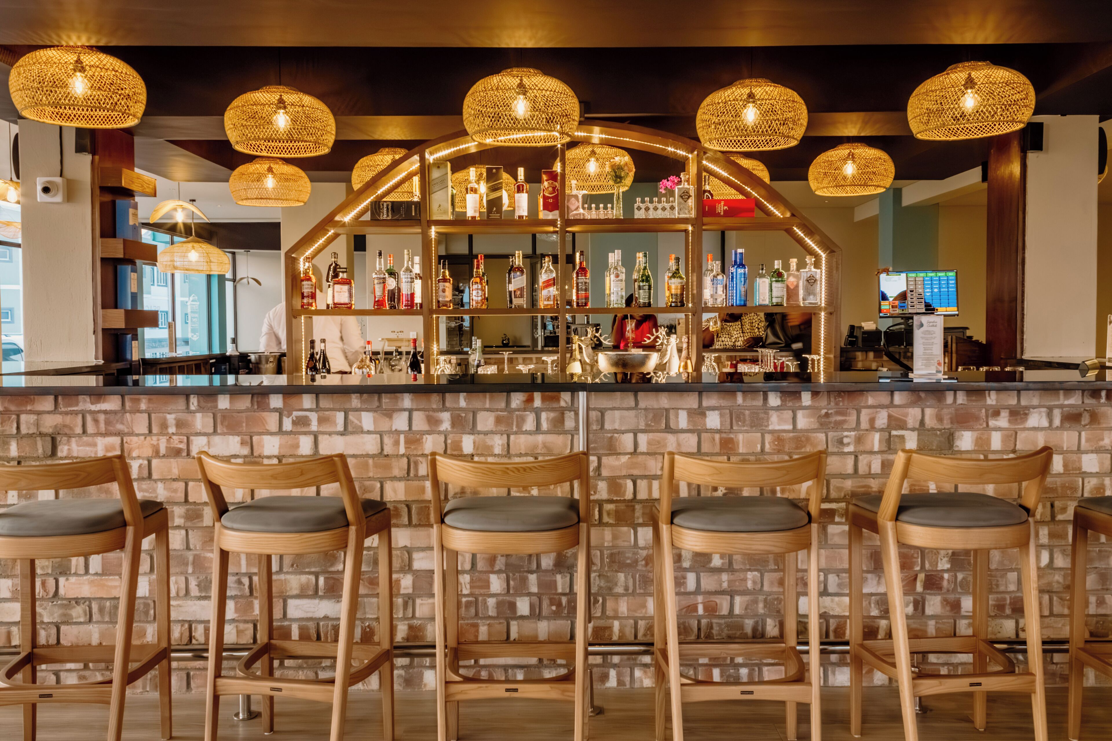 Uma área de bar com parede de tijolos, banquetas de madeira e variedade de garrafas e copos em prateleiras.