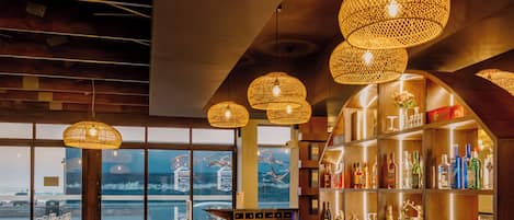 Uma área de bar com um balcão comprido, várias luminárias de vime pendentes e um display de vinhos e bebidas.