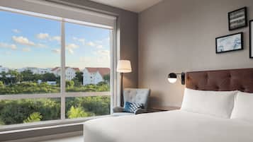 Quarto de hotel com uma janela grande, uma cama, uma cadeira e uma mesinha de cabeceira com abajur.