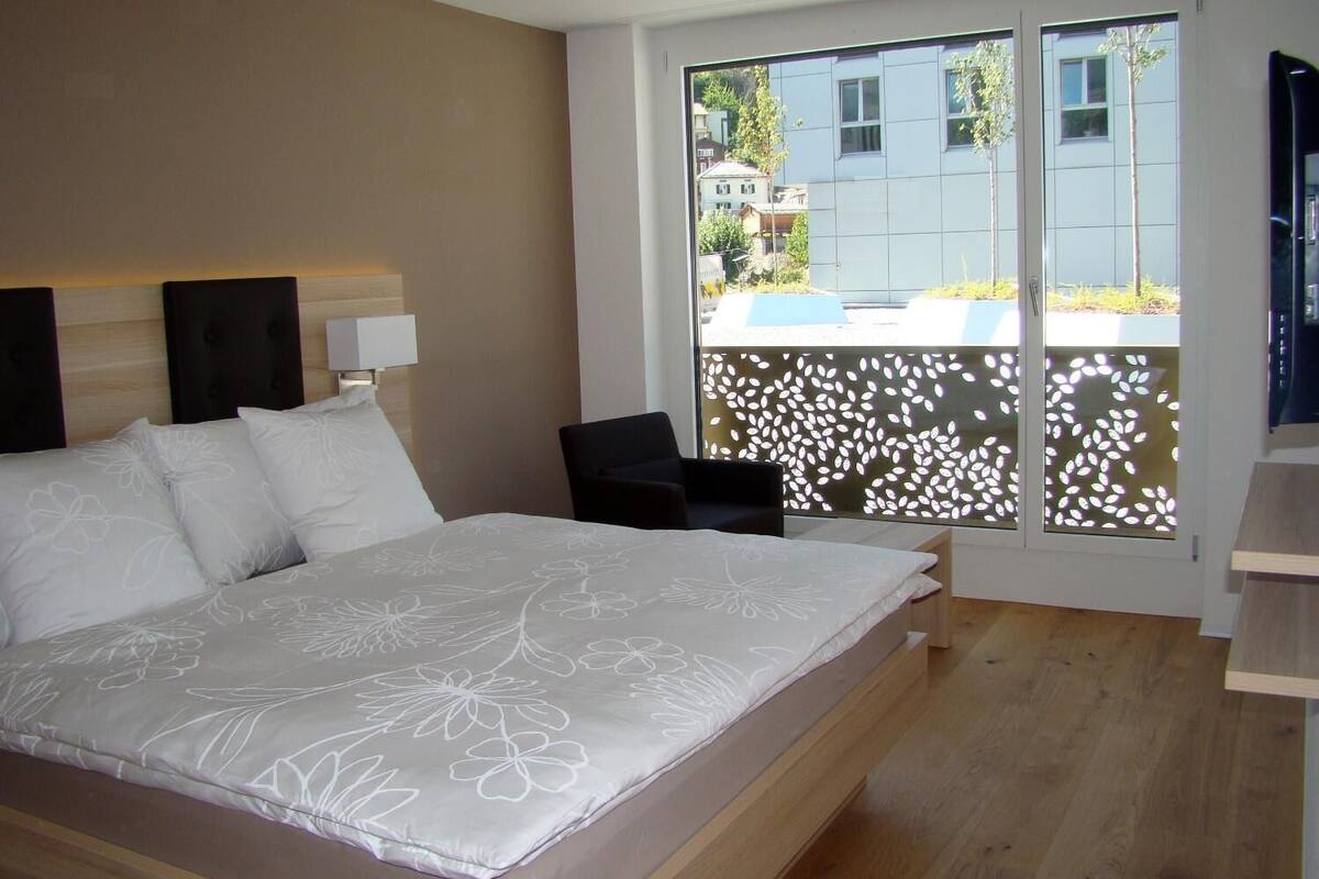 Quarto com uma cama grande, uma cadeira, uma escrivaninha e uma janela com vista para edifícios.