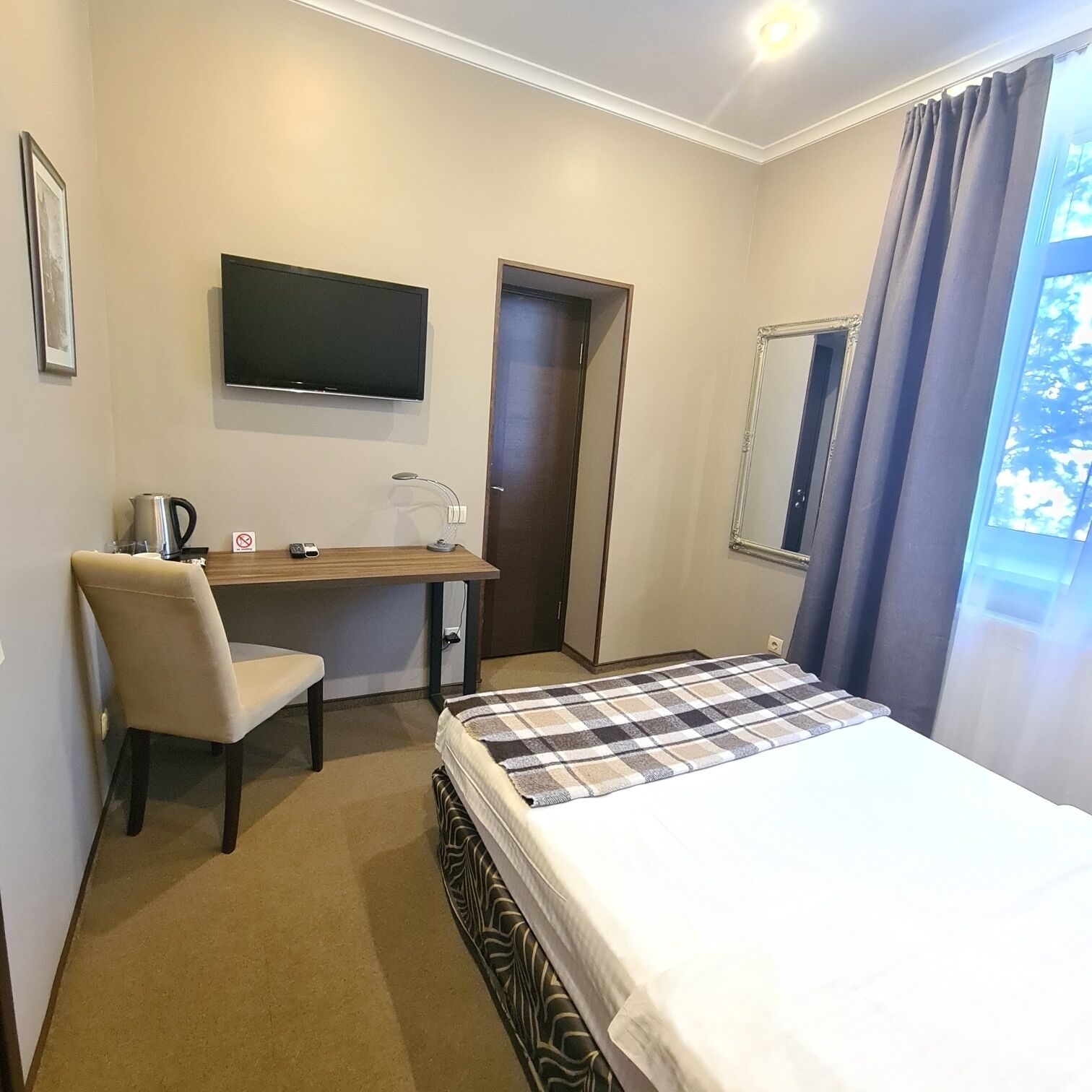 Quarto de hotel com uma cama, uma mesa com cadeira, televisão, porta e janela com cortinas.