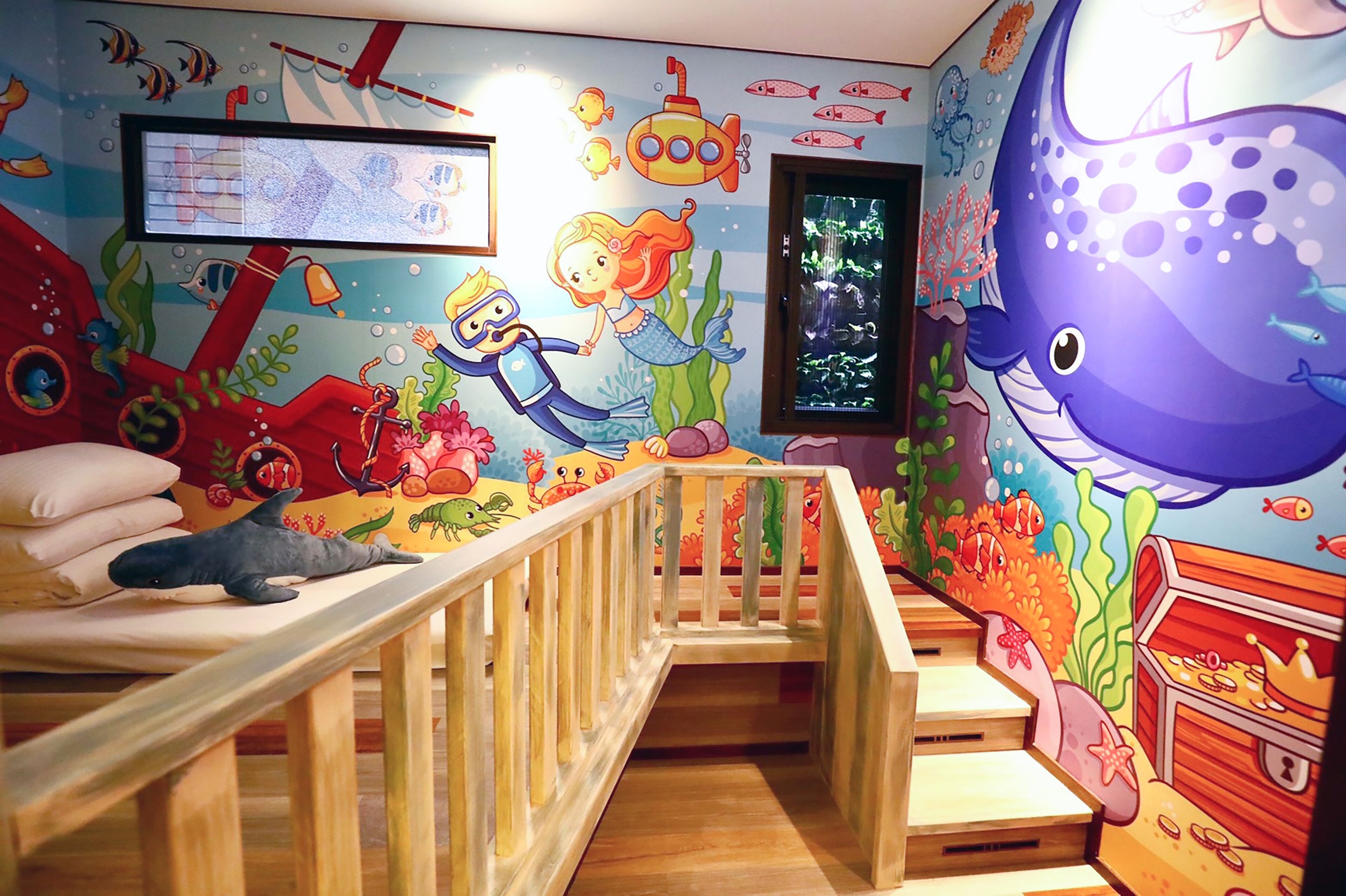 Uma sala de brinquedos infantil com escada de madeira, uma cama e um mural na parede com tema de fundo marinho.