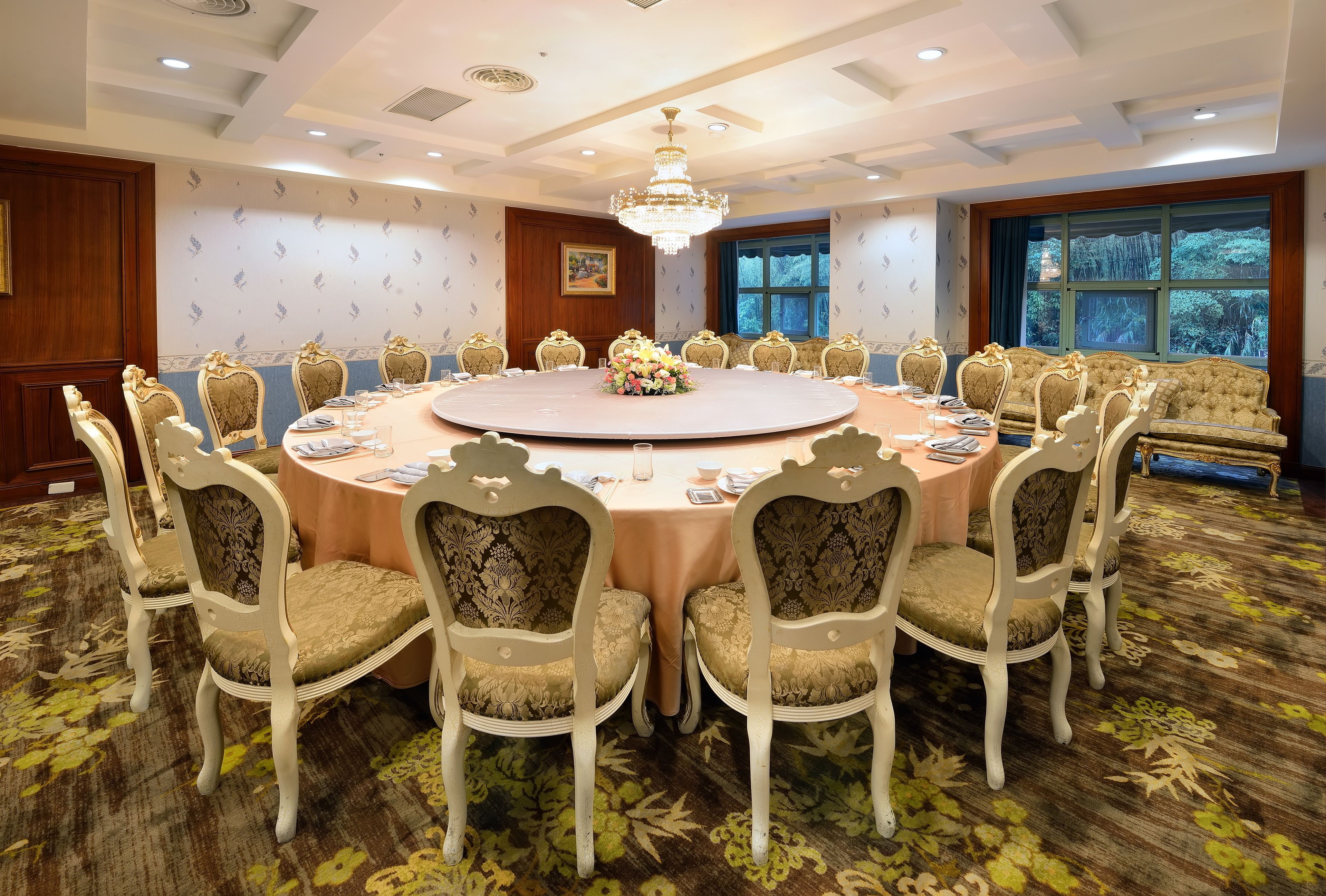 Uma sala de conferências com uma mesa redonda, cadeiras ornamentadas, um candelabro e uma grande janela com vista.