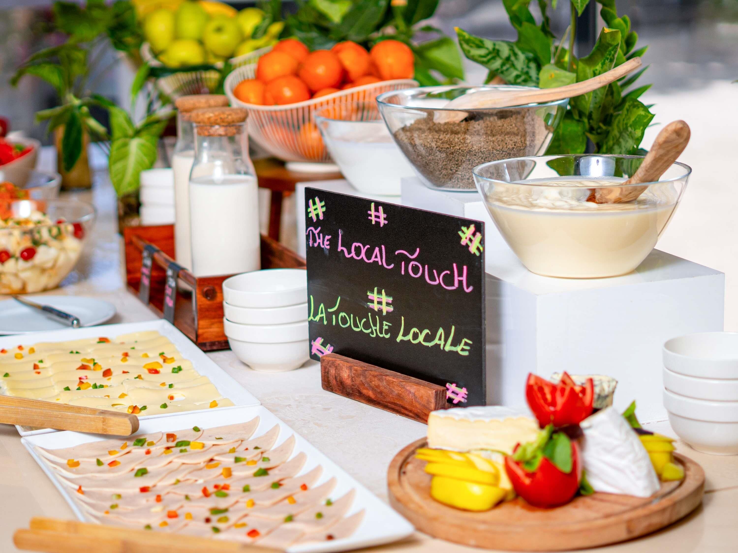 Uma mesa de buffet com diversos alimentos, um letreiro com a inscrição 'Local Touch' e uma tábua de madeira com queijo e tomates.
