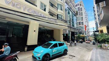 Hotel com entrada destacada, um carro azul estacionado na frente e uma passarela para pedestres.