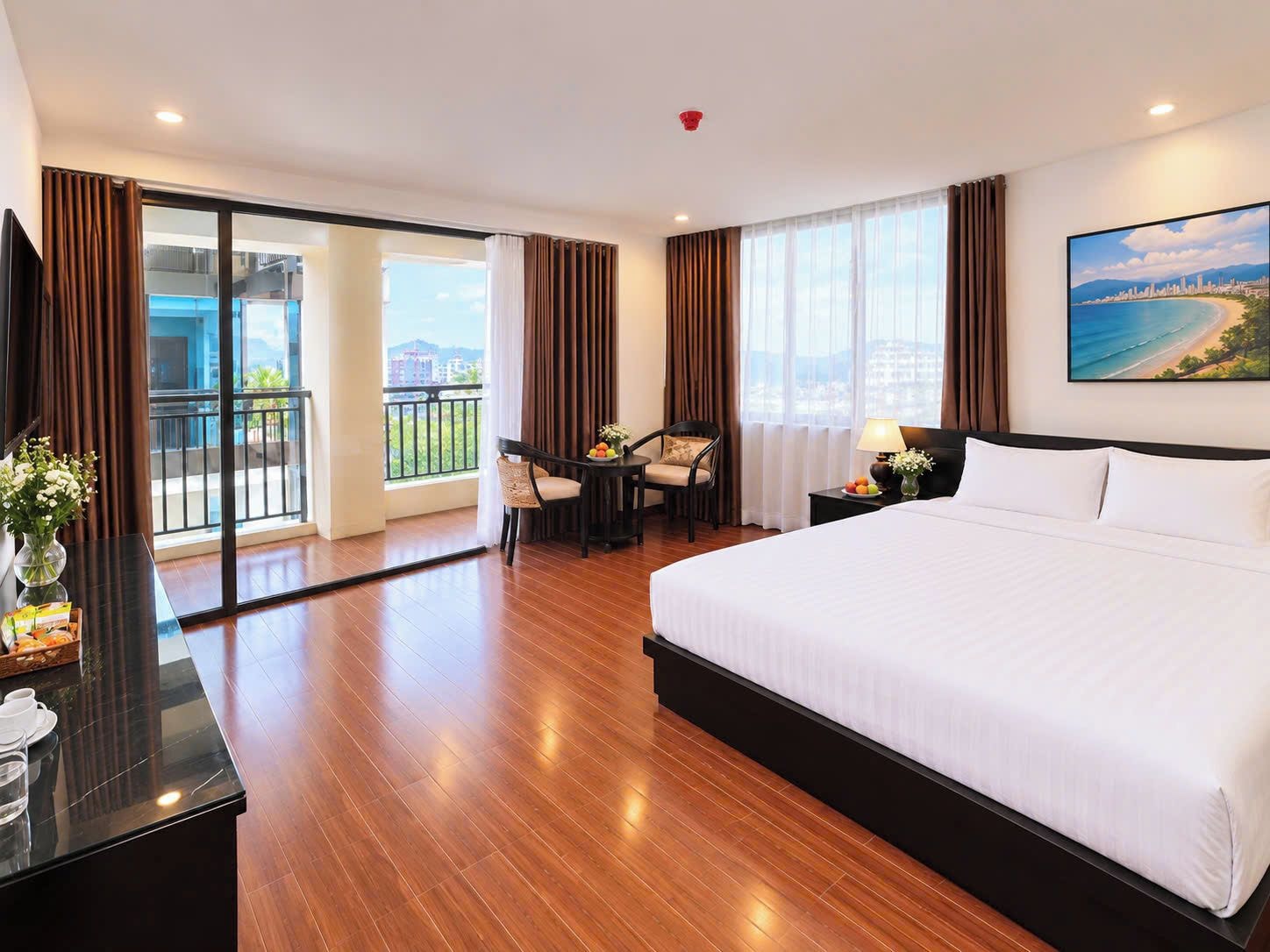 Quarto de hotel com uma cama grande, uma mesa de jantar com cadeiras, uma mesinha com abajur e vista para a cidade através de amplas janelas.
