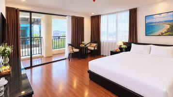 Quarto de hotel com uma cama grande, uma mesa de jantar com cadeiras, uma mesinha com abajur e vista para a cidade através de amplas janelas.
