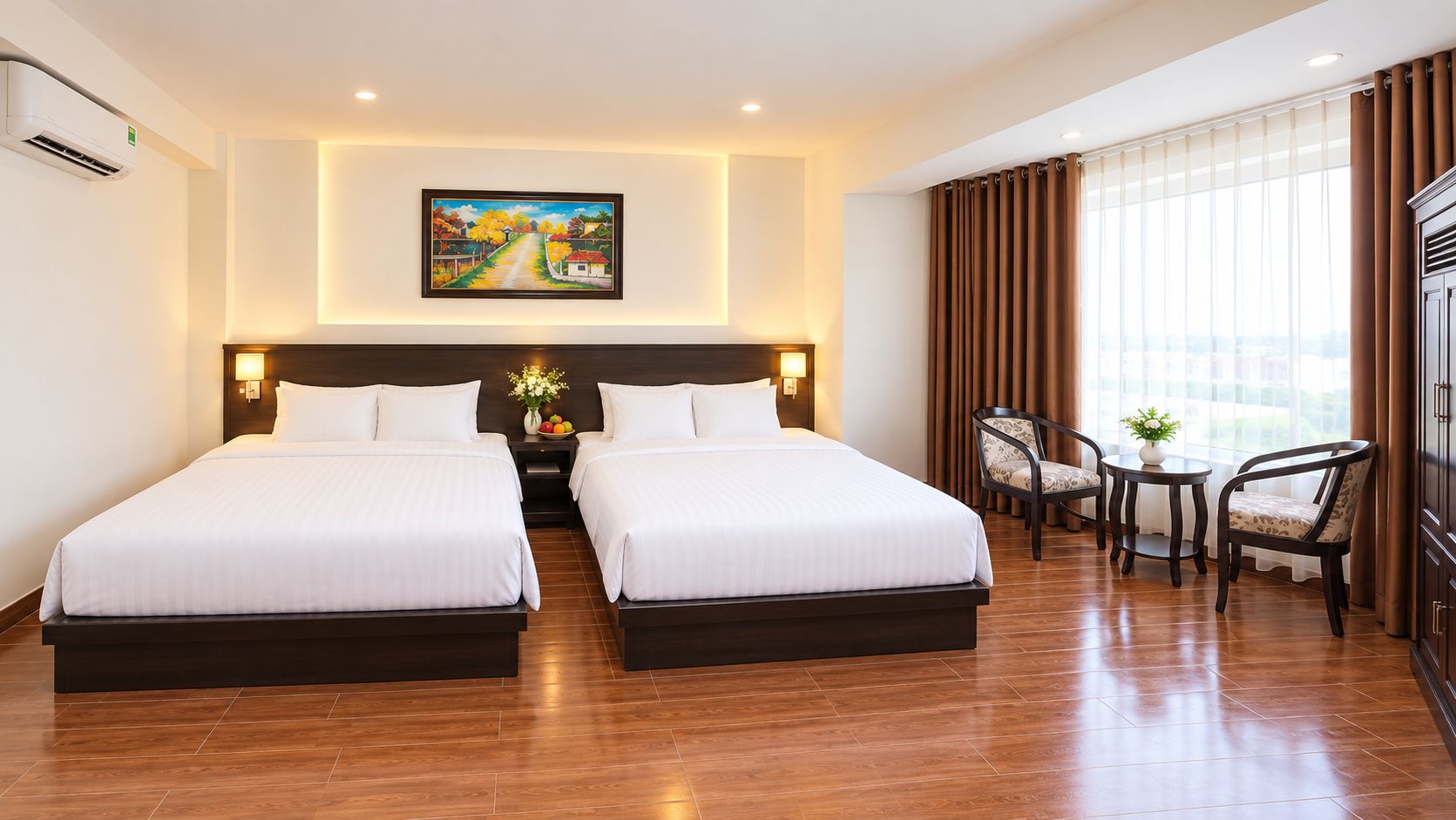 Quarto de hotel com duas camas, um quadro na parede, uma mesinha com um vaso e vista da cidade por grandes janelas.