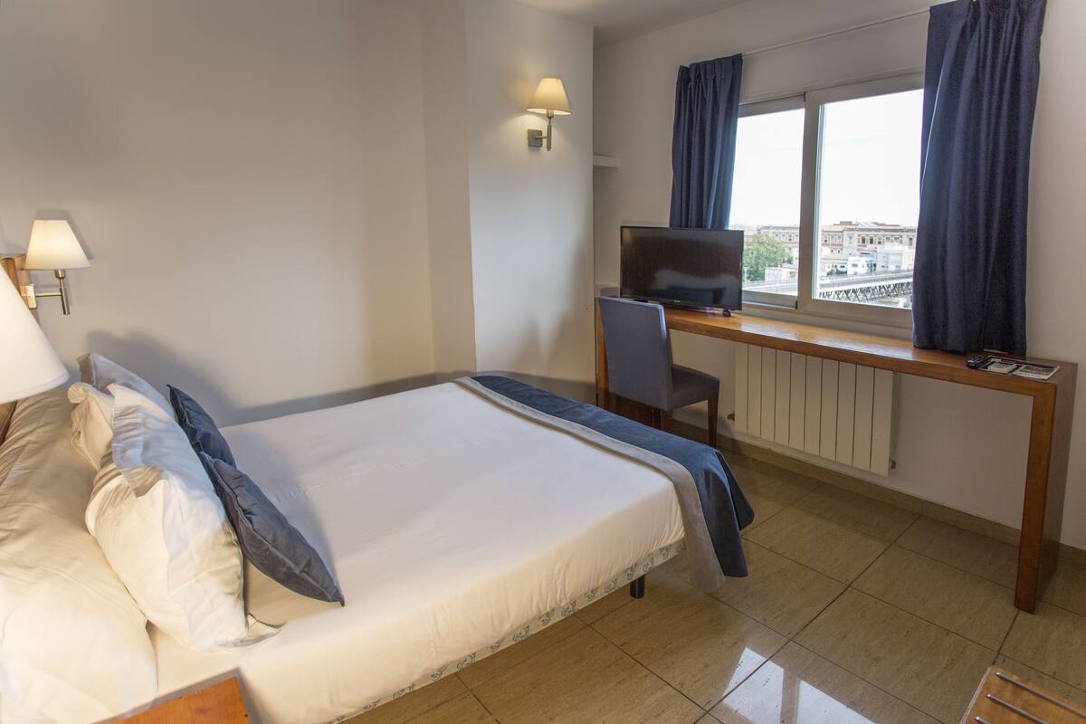 Quarto de hotel com cama, escrivaninha com computador, cadeira e janela com cortinas.