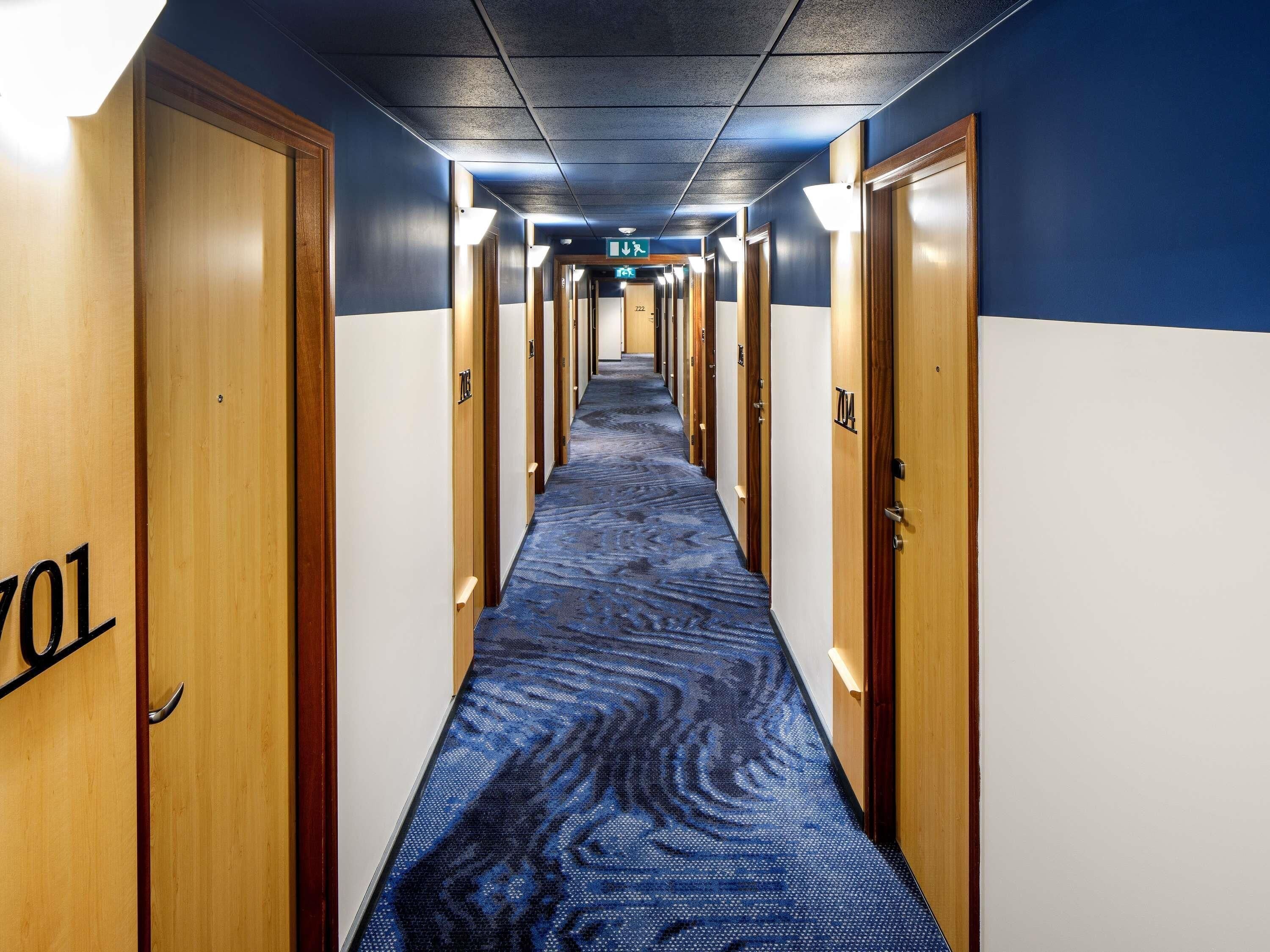 Corredor de hotel com portas de madeira, carpete azul e teto com iluminação embutida.
