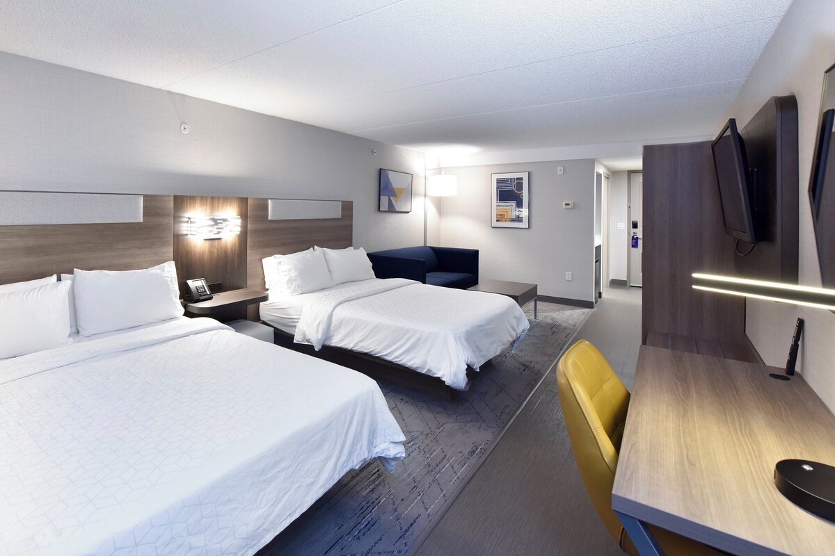 Quarto de hotel com duas camas, uma escrivaninha, uma cadeira, televisão e obras de arte nas paredes.