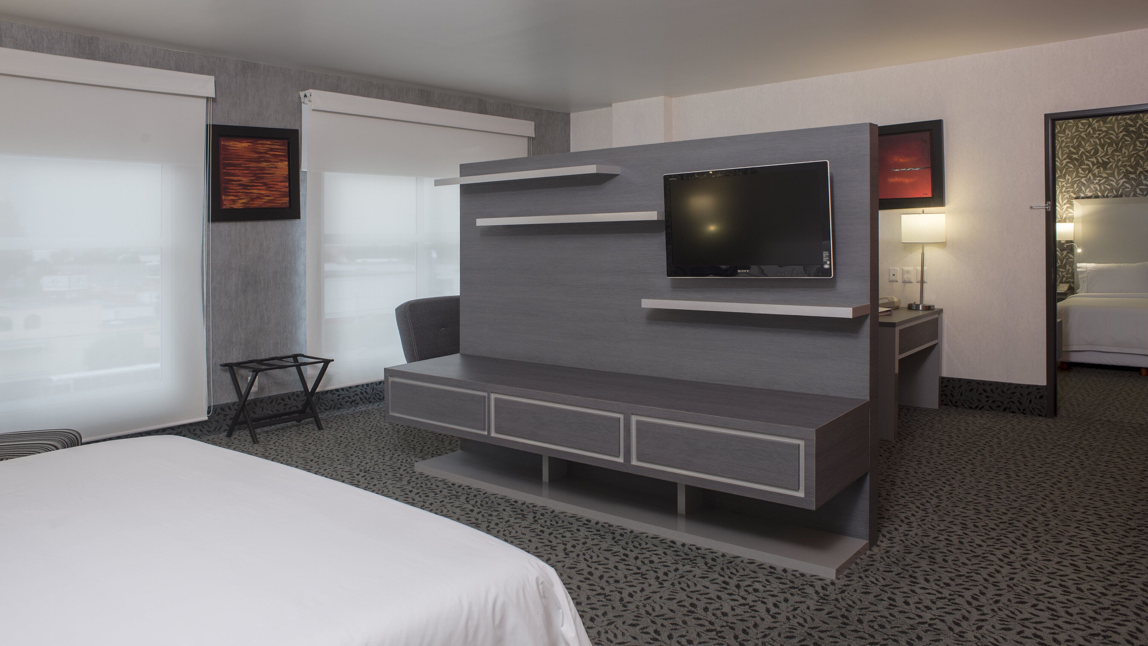 Quarto de hotel moderno com uma cama grande, uma televisão de tela plana presa a uma parede cinza, uma escrivaninha pequena com uma luminária e um tapete estampado.