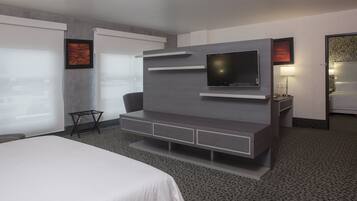 Quarto de hotel moderno com uma cama grande, uma televisão de tela plana presa a uma parede cinza, uma escrivaninha pequena com uma luminária e um tapete estampado.