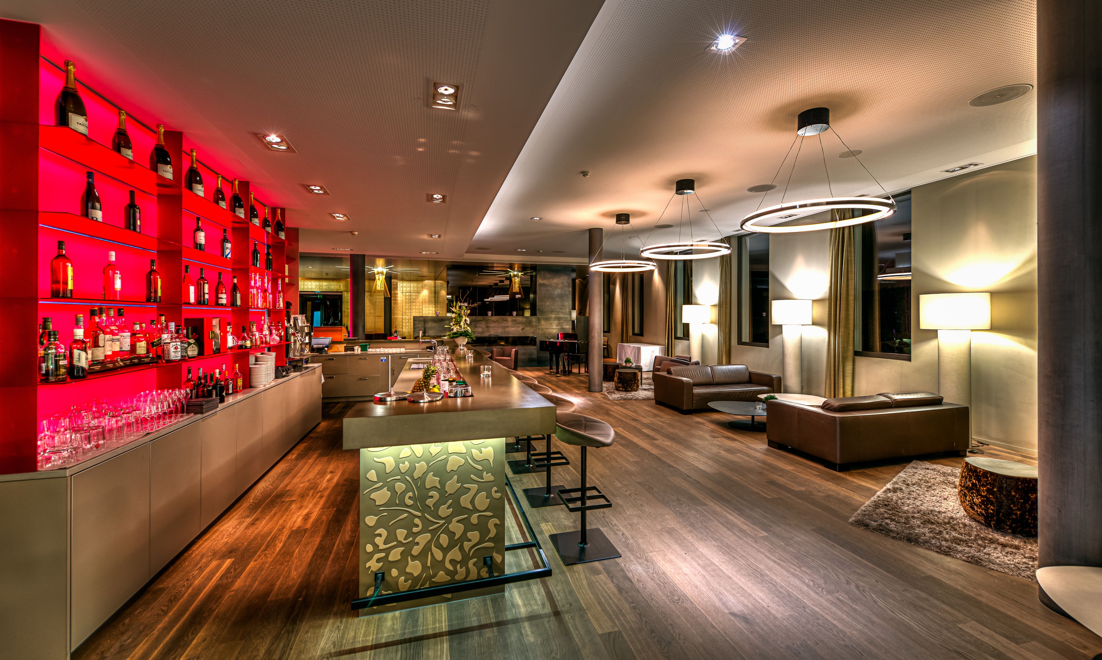 Uma área de bar moderna com uma adega iluminada em vermelho, um balcão de bar alongado e assentos, com um piano ao fundo.