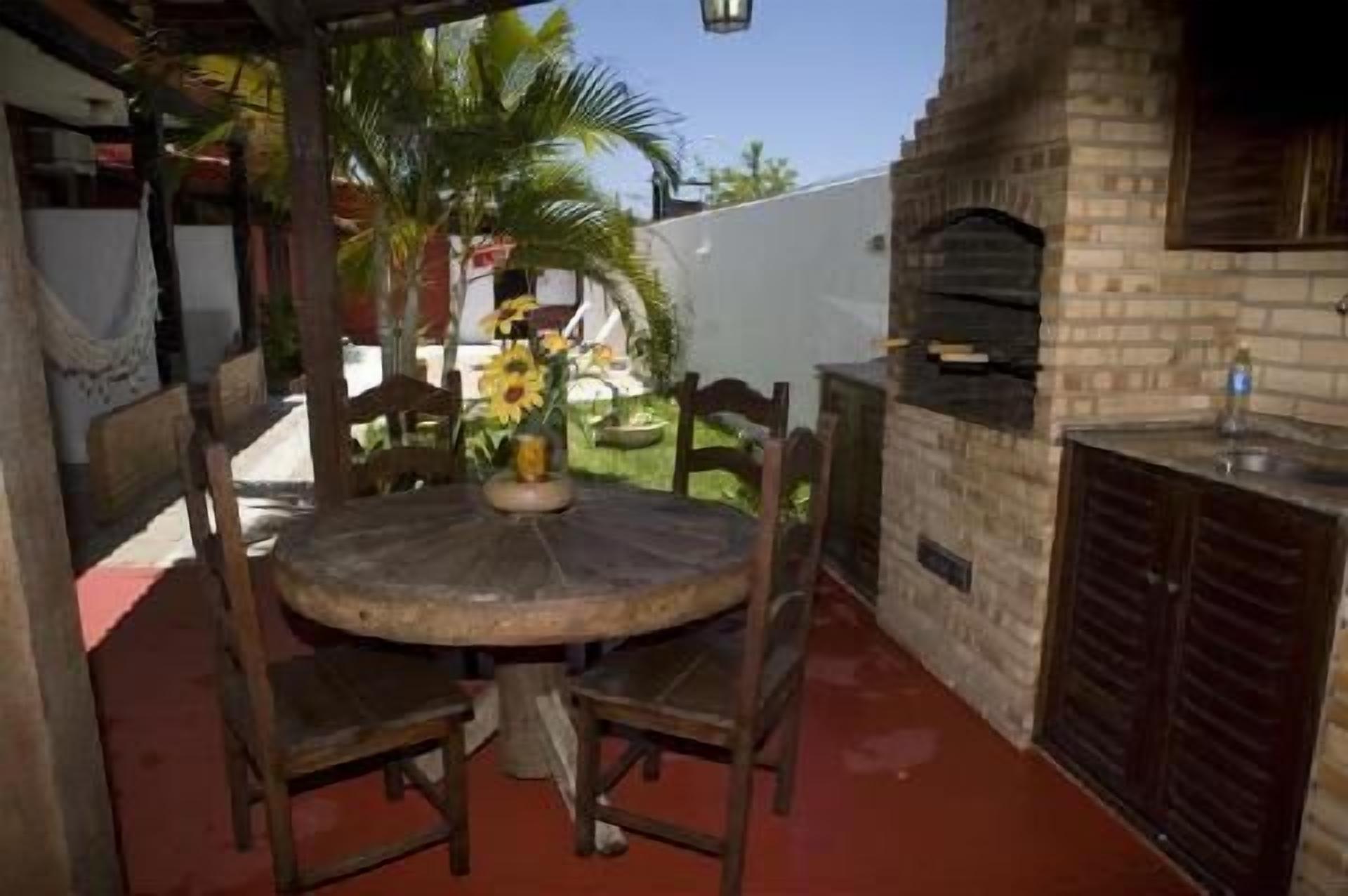 Um pátio externo com mesa redonda e cadeiras, um forno de tijolos e um pequeno jardim com girassóis.