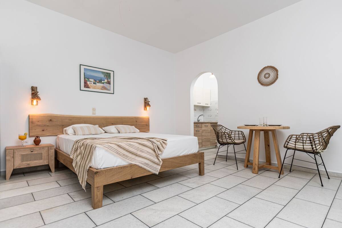 Quarto com cama de madeira, mesas de cabeceira, uma pequena mesa com cadeiras, luminária na parede e um quadro.