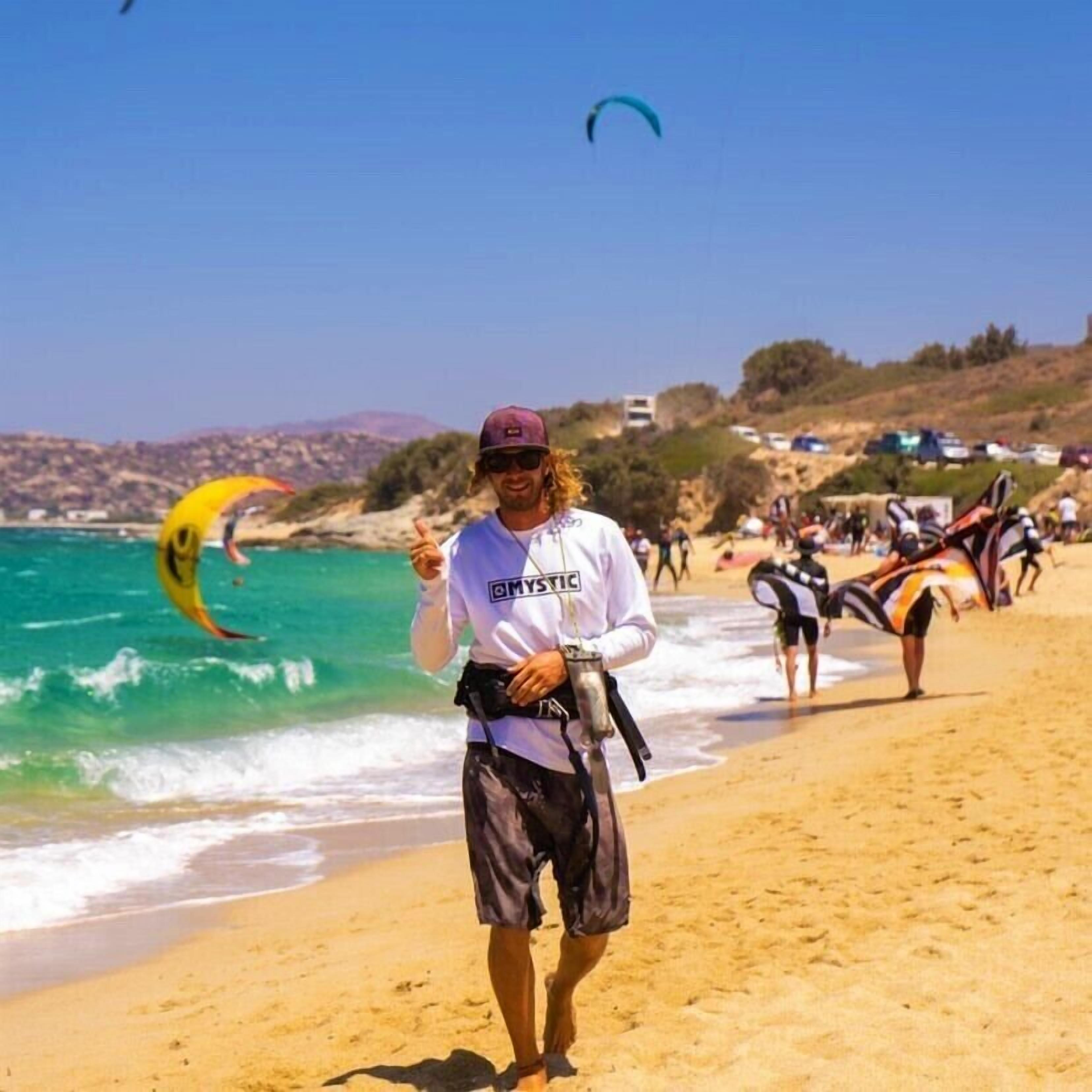 Uma praia com pessoas, incluindo alguém de camisa e boné branco, e um pipa amarelo.