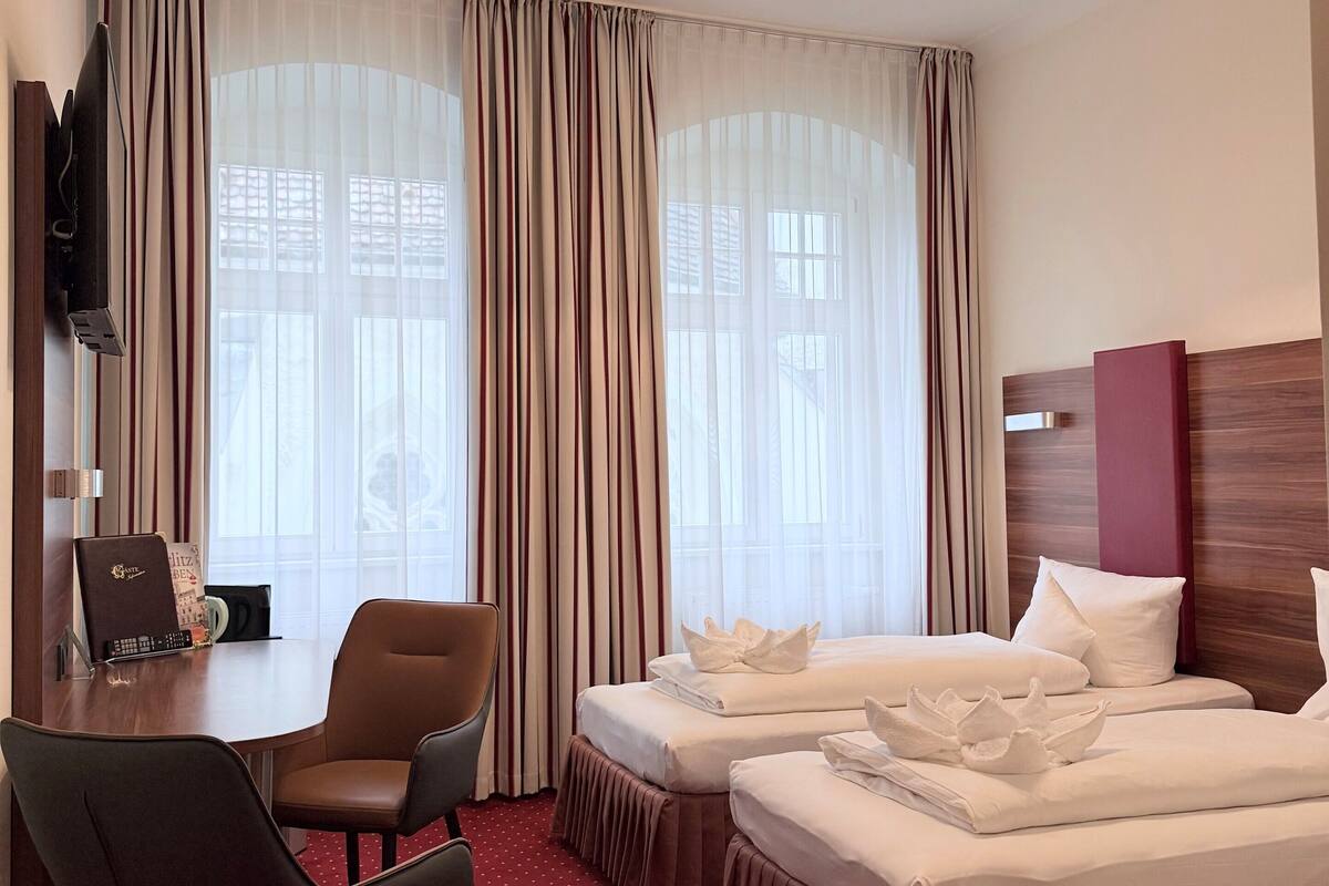 Quarto de hotel com duas camas, uma escrivaninha com cadeira, televisão e uma janela grande com cortinas.