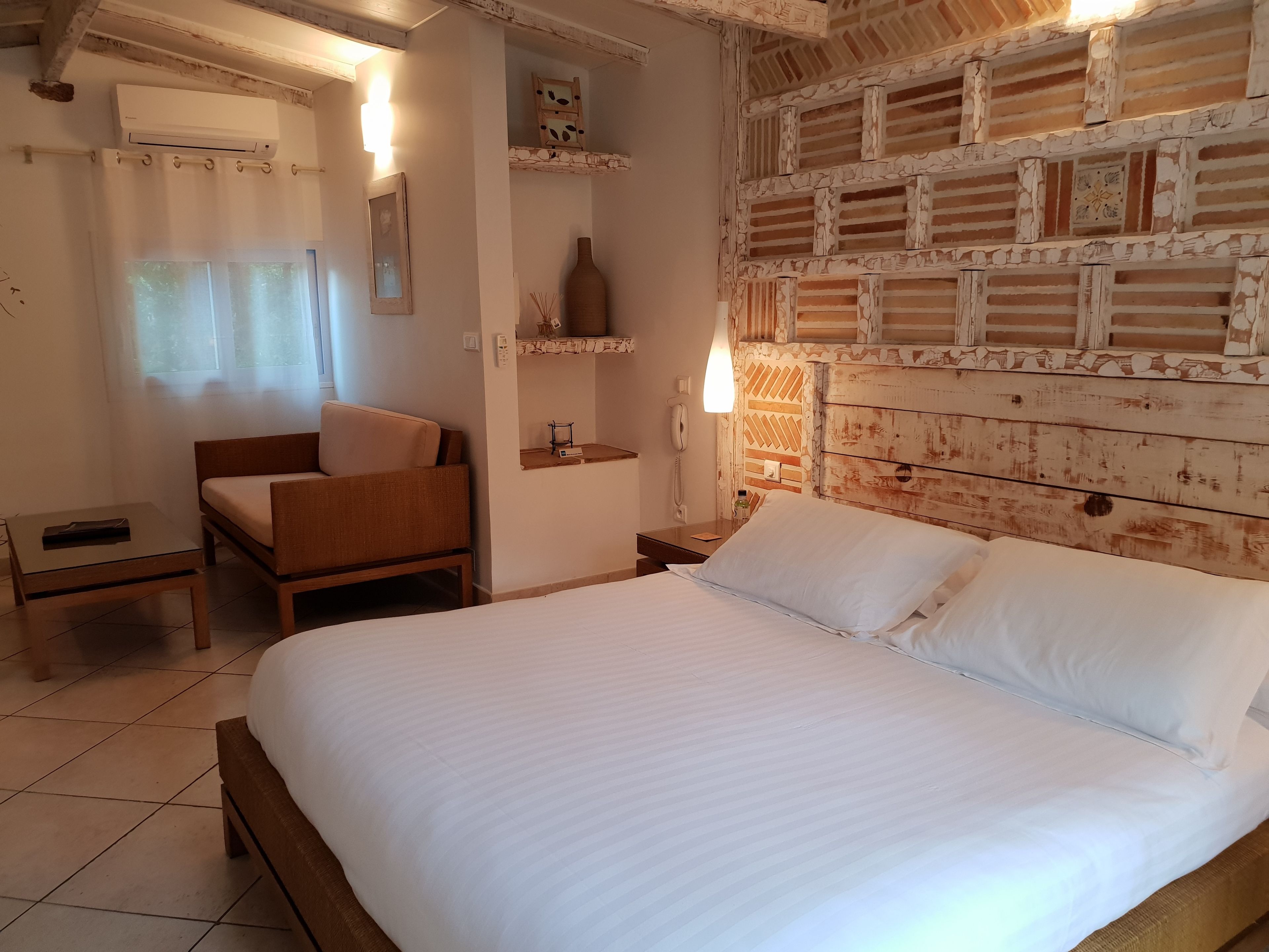 Quarto com uma cama grande, uma cadeira de madeira, uma mesa de cabeceira, um criado-mudo, um telefone fixado na parede e uma prateleira decorativa.