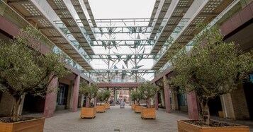 Uma galeria coberta com teto de vidro, ladeada por árvores e bancos de madeira.