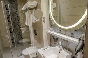 Uma casa de banho moderna com cabine de duche em vidro, lavatório branco, espelho com moldura iluminada e toalheiro com toalhas dobradas.