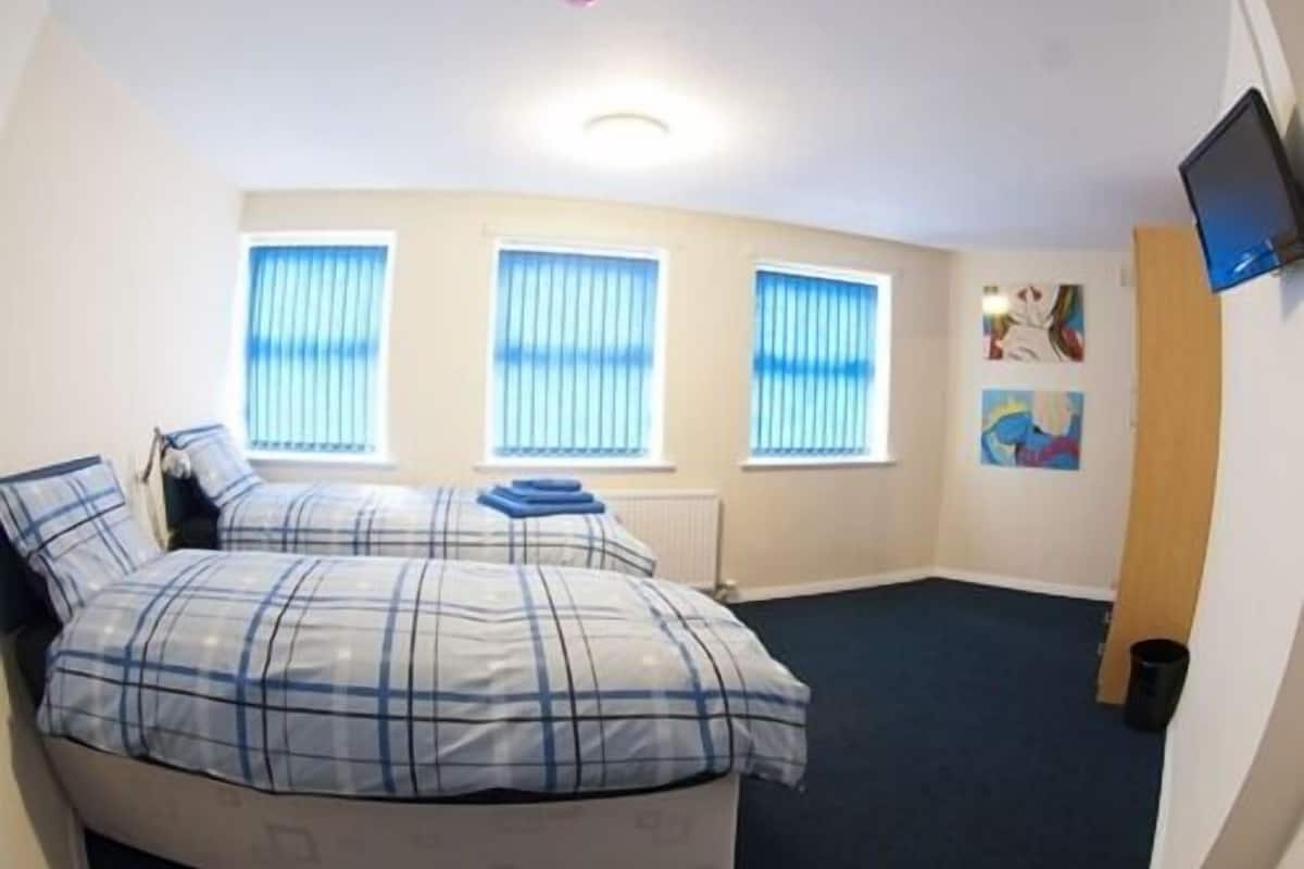 Quarto com cama, três janelas, televisão presa à parede e obras de arte.