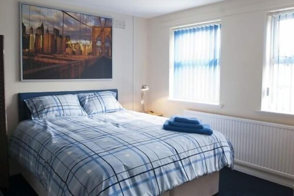 Quarto com cama, janela com cortinas, radiador e um quadro na parede.