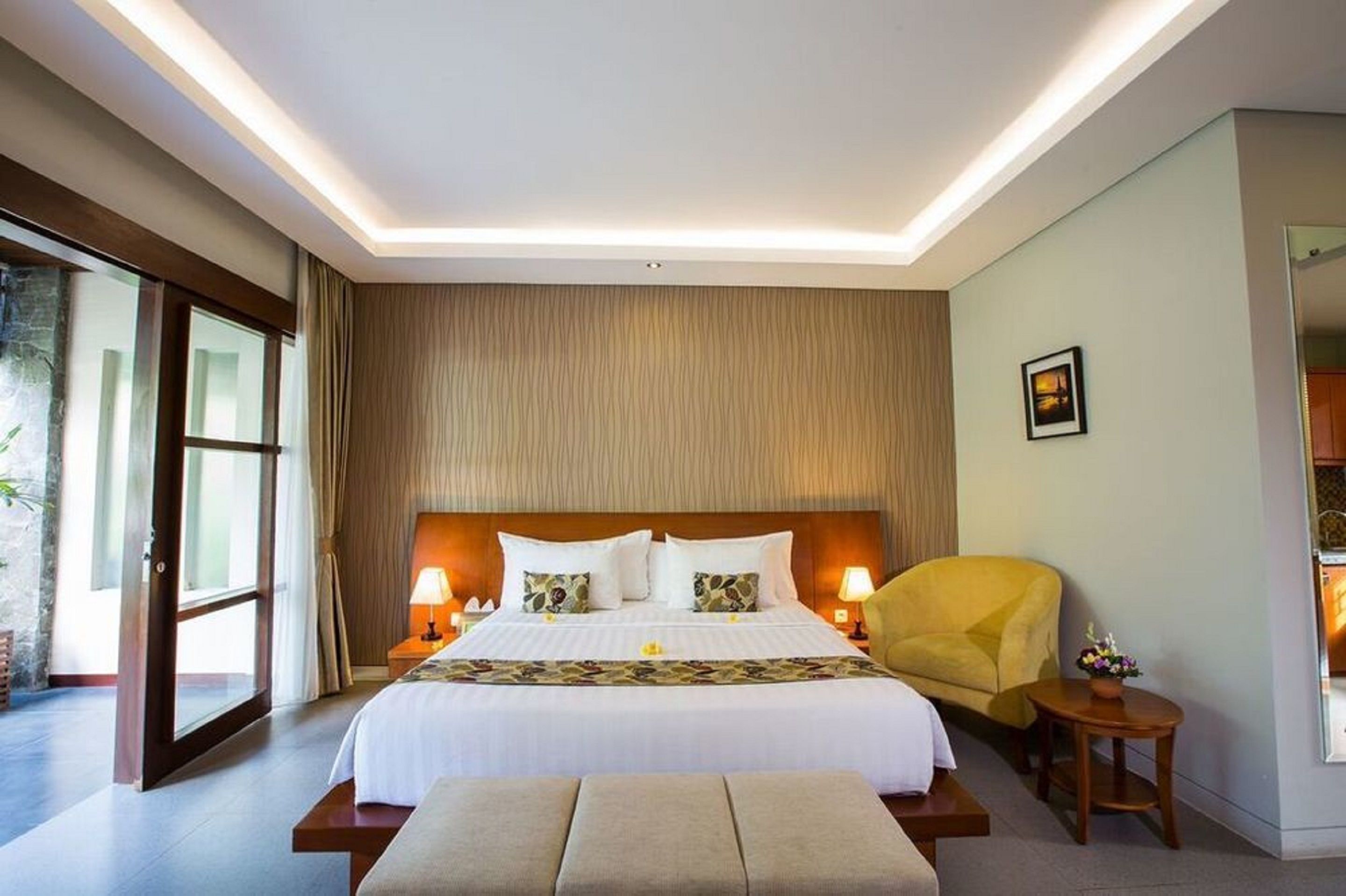 Quarto de hotel moderno com uma cama grande, abajures, uma poltrona amarela e uma mesinha com flores.