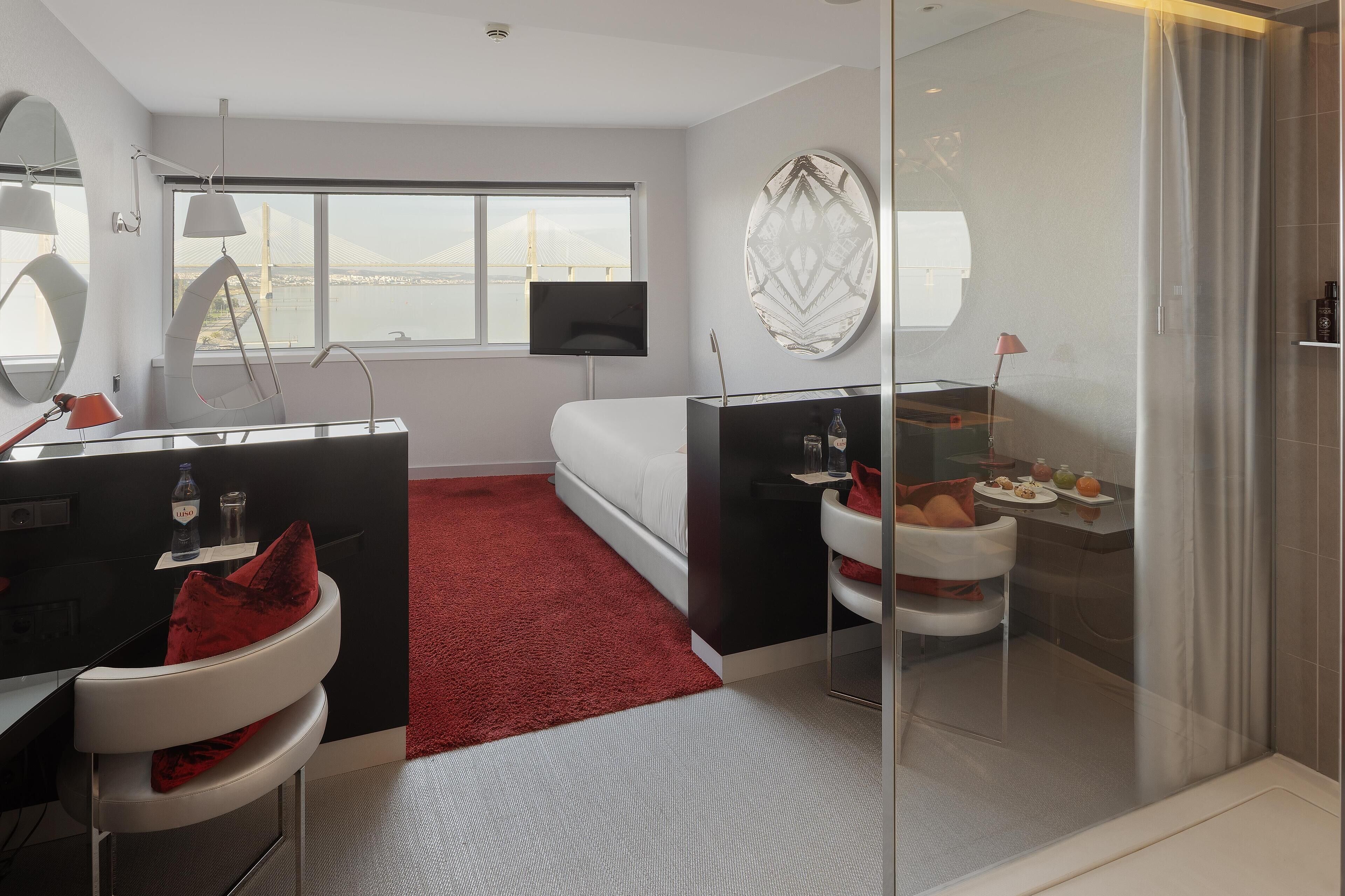 Quarto de hotel moderno com cama, escrivaninha com computador, box de vidro para o chuveiro e vista para uma ponte.