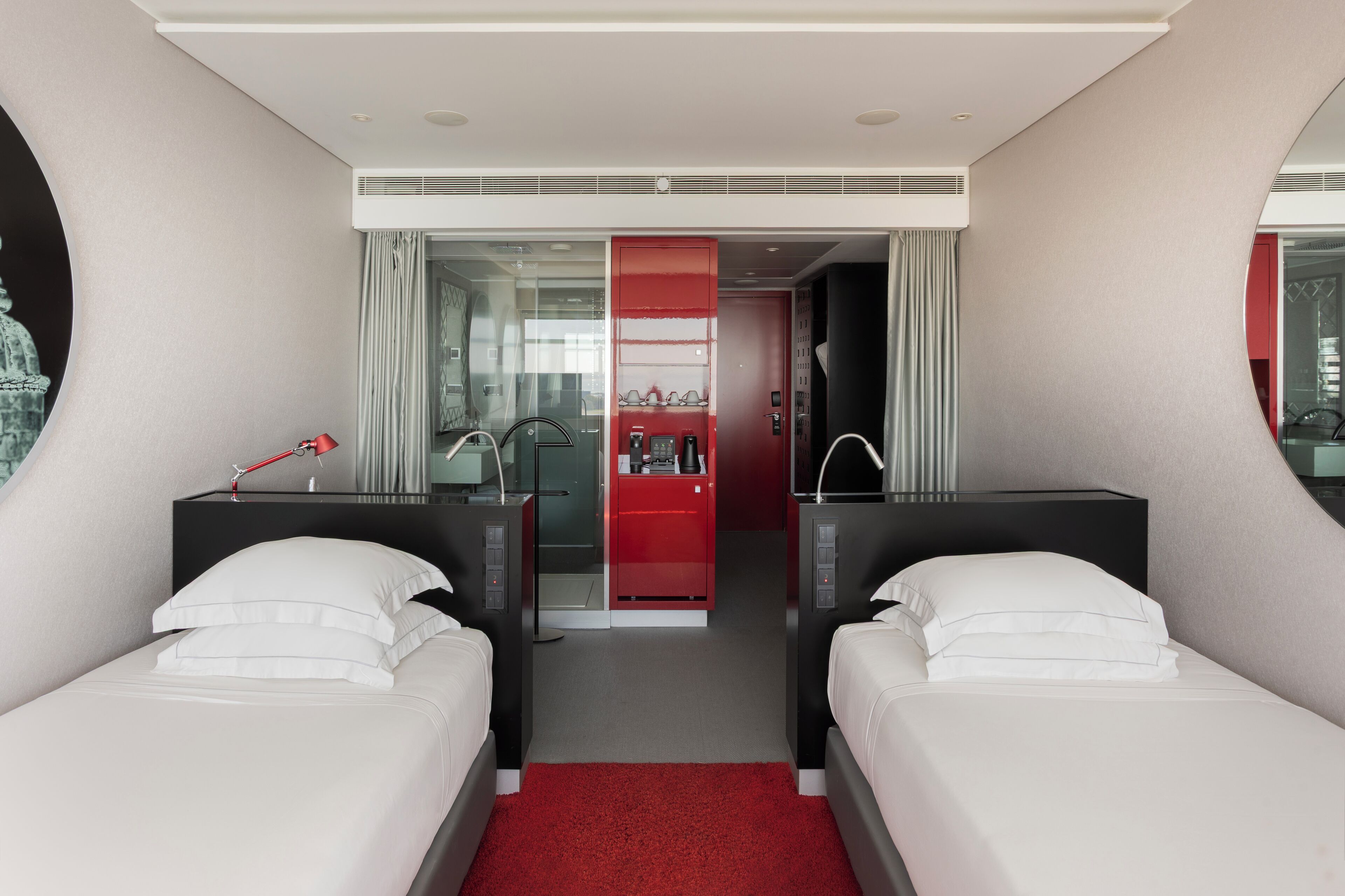Quarto de hotel com duas camas, um minibar vermelho e um painel de vidro separando o quarto do corredor.