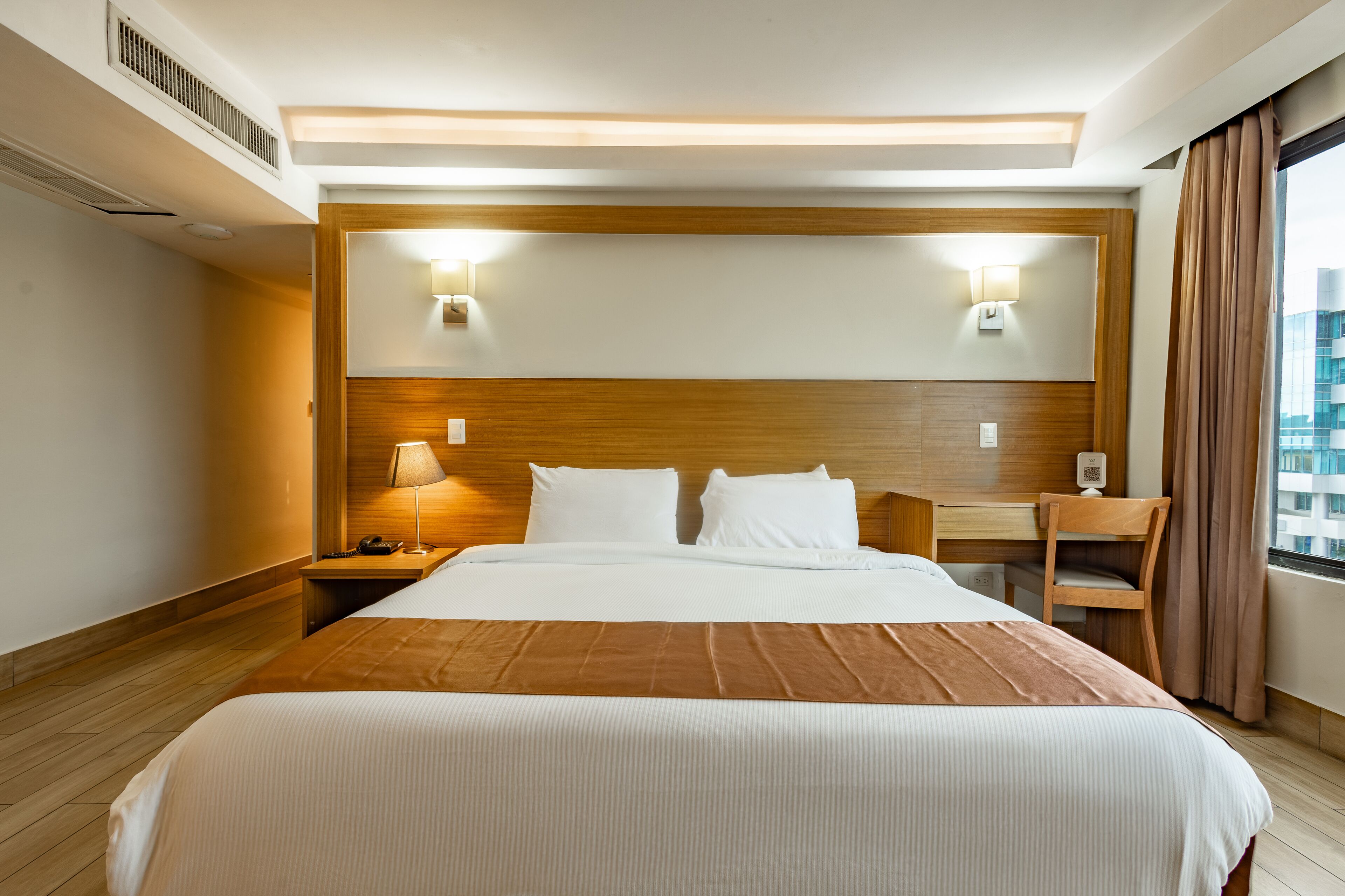 Quarto de hotel com uma cama grande, uma mesa de trabalho, duas luminárias na parede e vista da cidade pela janela.
