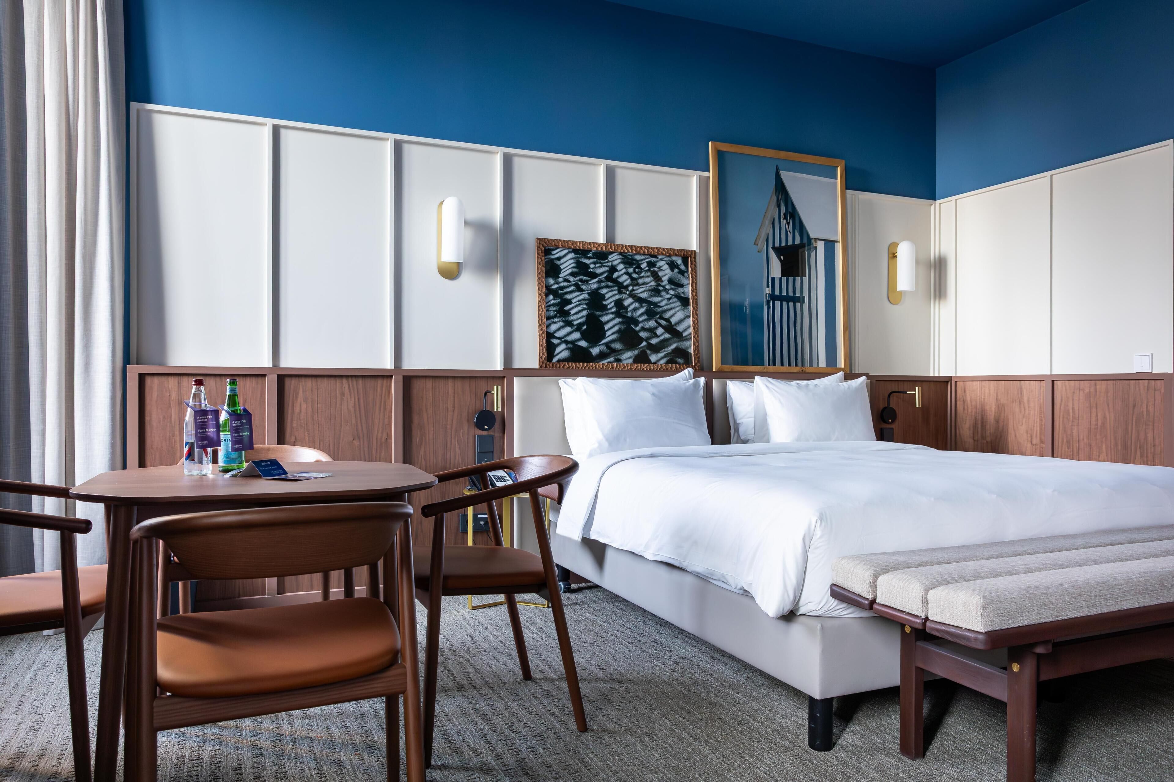 Quarto de hotel com uma cama, uma escrivaninha, duas cadeiras, um banco, um espelho e uma obra de arte emoldurada na parede.