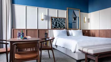 Quarto de hotel com uma cama, uma escrivaninha, duas cadeiras, um banco, um espelho e uma obra de arte emoldurada na parede.