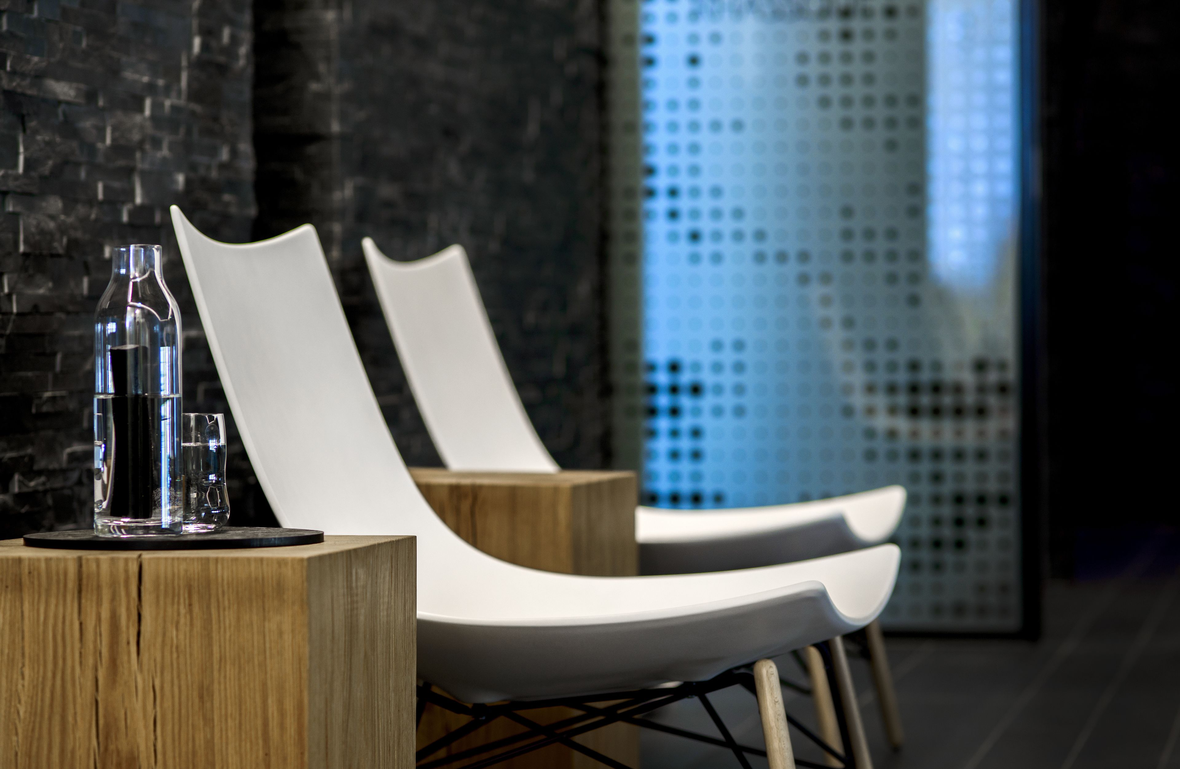 Uma área de estar moderna com cadeiras brancas e uma mesa lateral de madeira, uma garrafa de vidro e uma parede metálica com design perfurado.