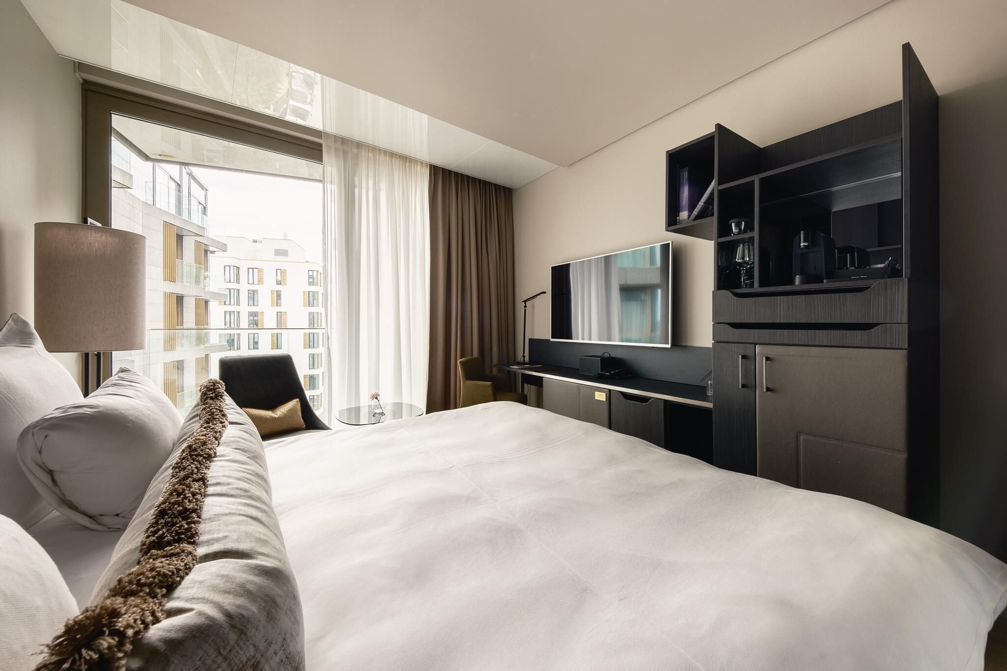 Quarto de hotel moderno com uma cama grande, televisão, secretária e vista para edifícios pela janela.