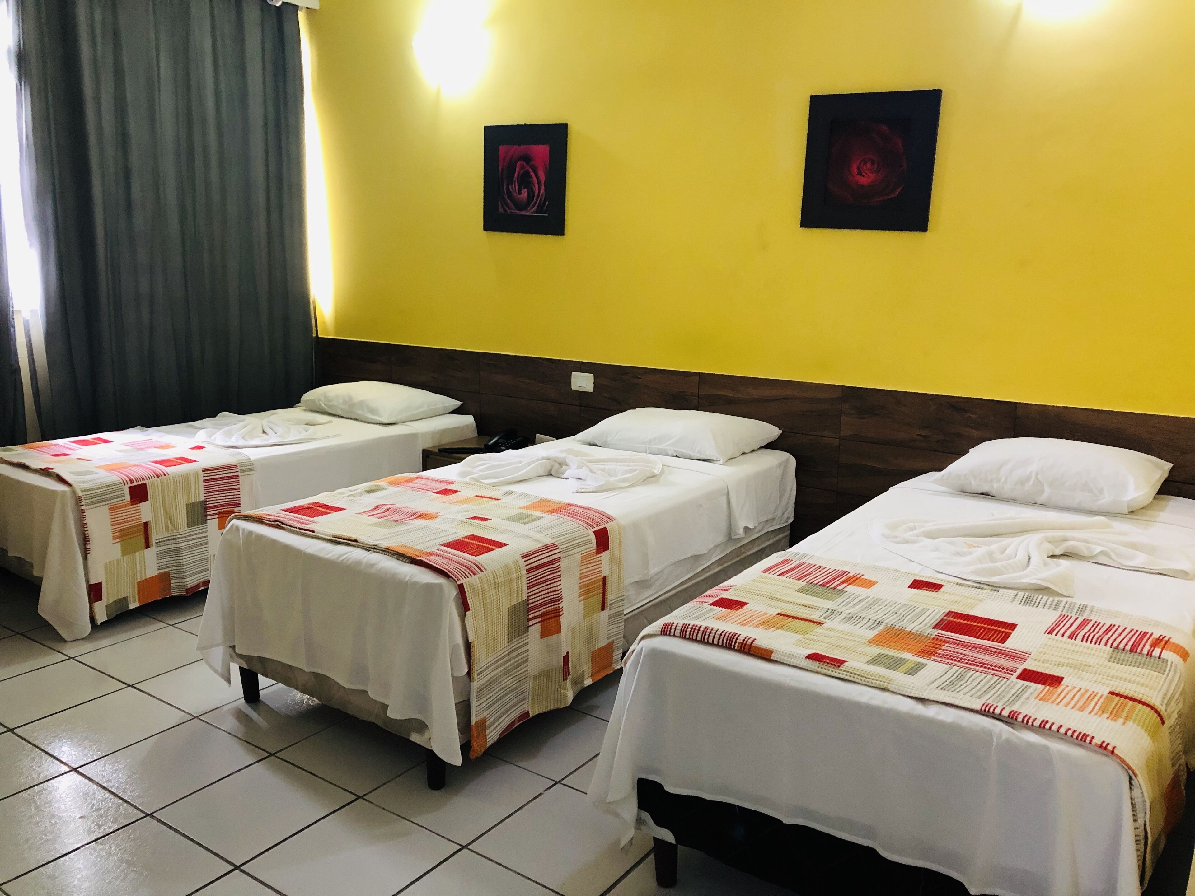 Quarto de hotel com três camas de solteiro, cada uma com um cobreleite branco e um edredom colorido. O quarto tem uma parede amarela e uma cabeceira de madeira.