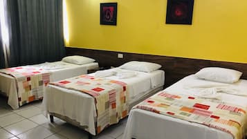 Quarto de hotel com três camas de solteiro, cada uma com um cobreleite branco e um edredom colorido. O quarto tem uma parede amarela e uma cabeceira de madeira.