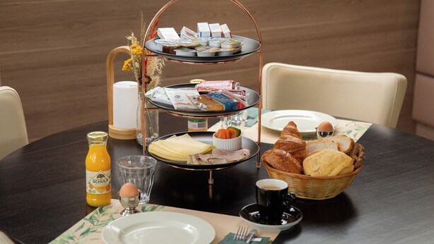 Mesa de café da manhã com bandeja escalonada, pão, doces, uma garrafa de suco de laranja e uma xícara de café.