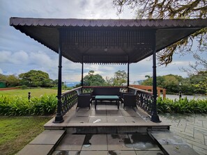 Área de estar coberta ao ar livre, com um banco e uma mesa, cercada por guarda-corpo de metal e vista para um lago e árvores.