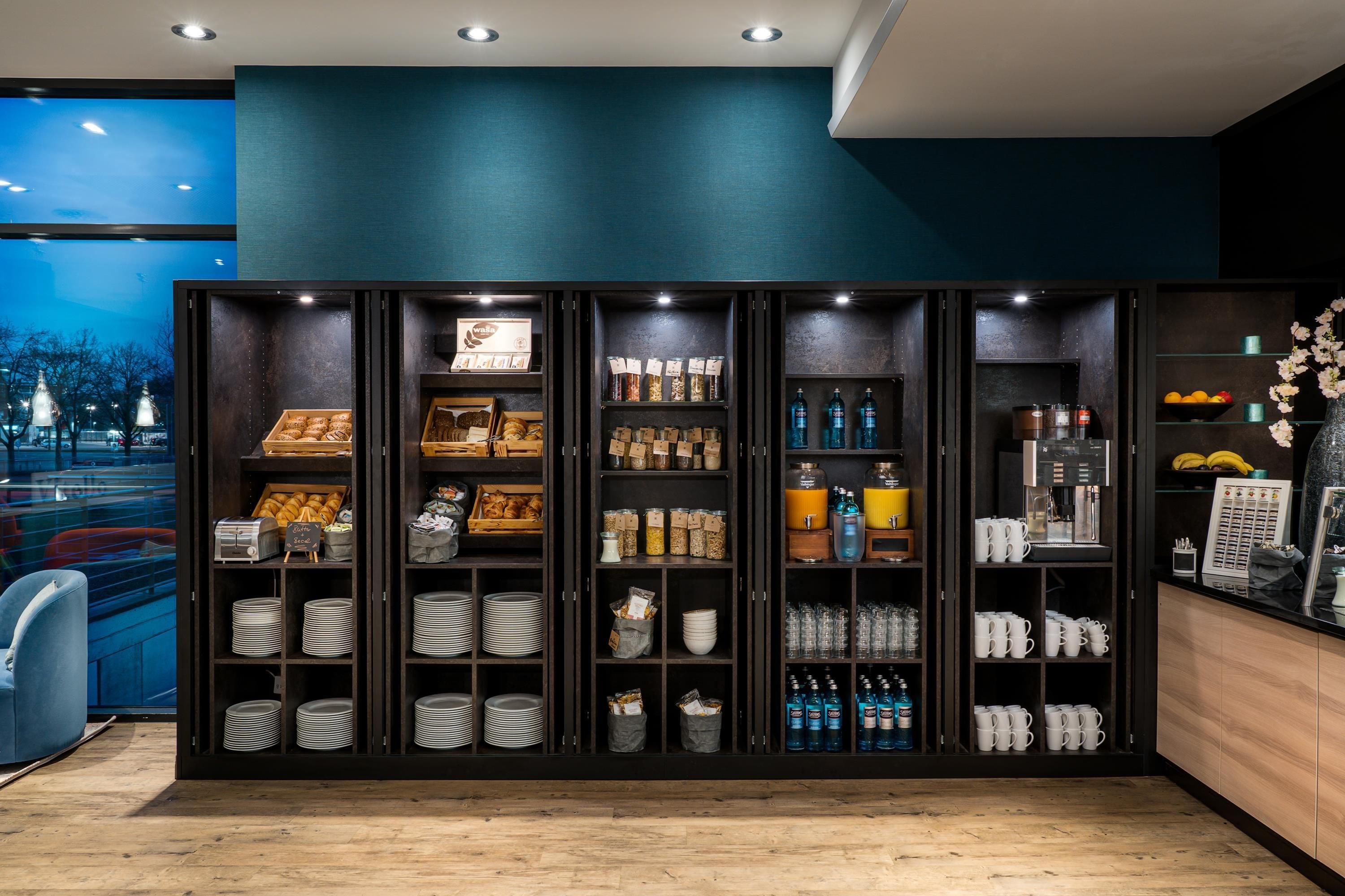 Interior de café com balcão de madeira escura, prateleiras repletas de produtos alimentícios e máquina de café.