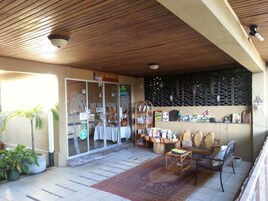 Área externa coberta com teto de madeira, entrada com porta de vidro, uma pequena mesa com uma cadeira, e uma exibição de livros e lembranças.