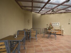 Uma sala de jantar com mesas e cadeiras de madeira, piso ladrilhado e teto alto com vigas de metal.