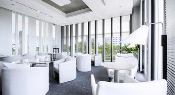 Quarto de hotel moderno com varanda, mobiliário branco e grandes janelas com vista para a cidade.