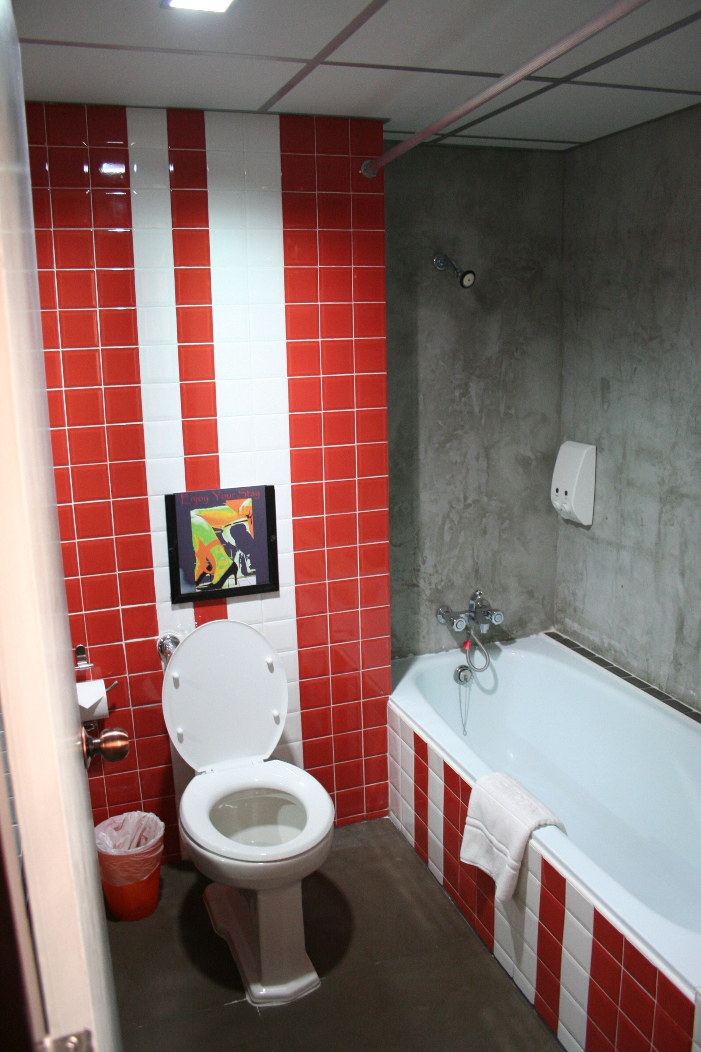 Uma casa de banho com sanita branca, paredes com azulejos vermelhos e brancos, uma banheira e um secador de mãos.