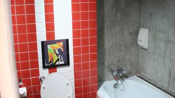 Uma casa de banho com sanita branca, paredes com azulejos vermelhos e brancos, uma banheira e um secador de mãos.