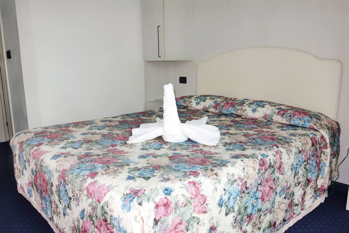 Cama bem arrumada com colcha florida e toalha branca dobrada ao centro.